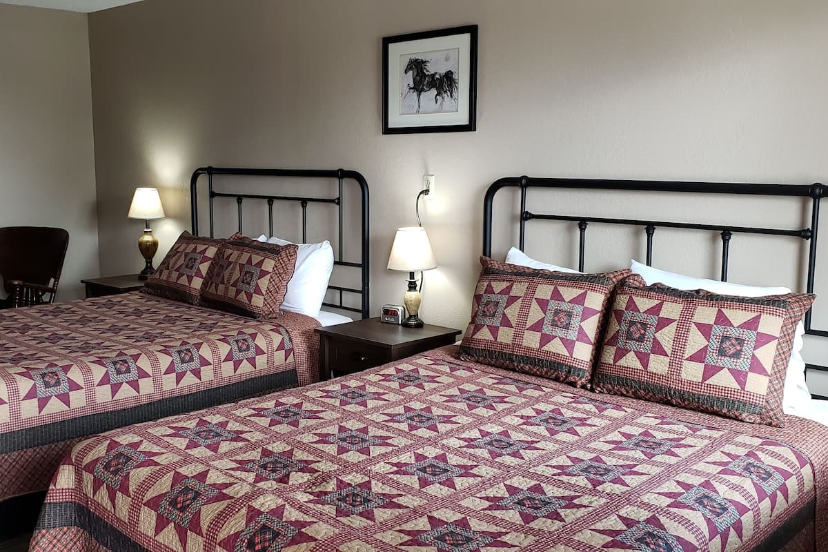 Quarto de hotel com duas camas, cada uma com colcha estampada e almofadas combinando. Possui mesas de cabeceira com luminárias, uma cadeira e obras de arte nas paredes.