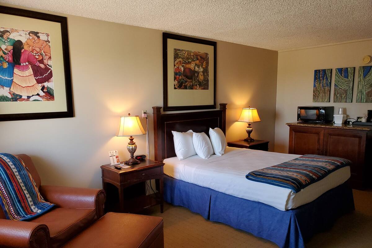 Quarto de hotel com cama, duas luminárias de cabeceira, mesa de cabeceira de madeira, poltrona de couro marrom, quadro pendurado na parede e área de cozinha compacta.