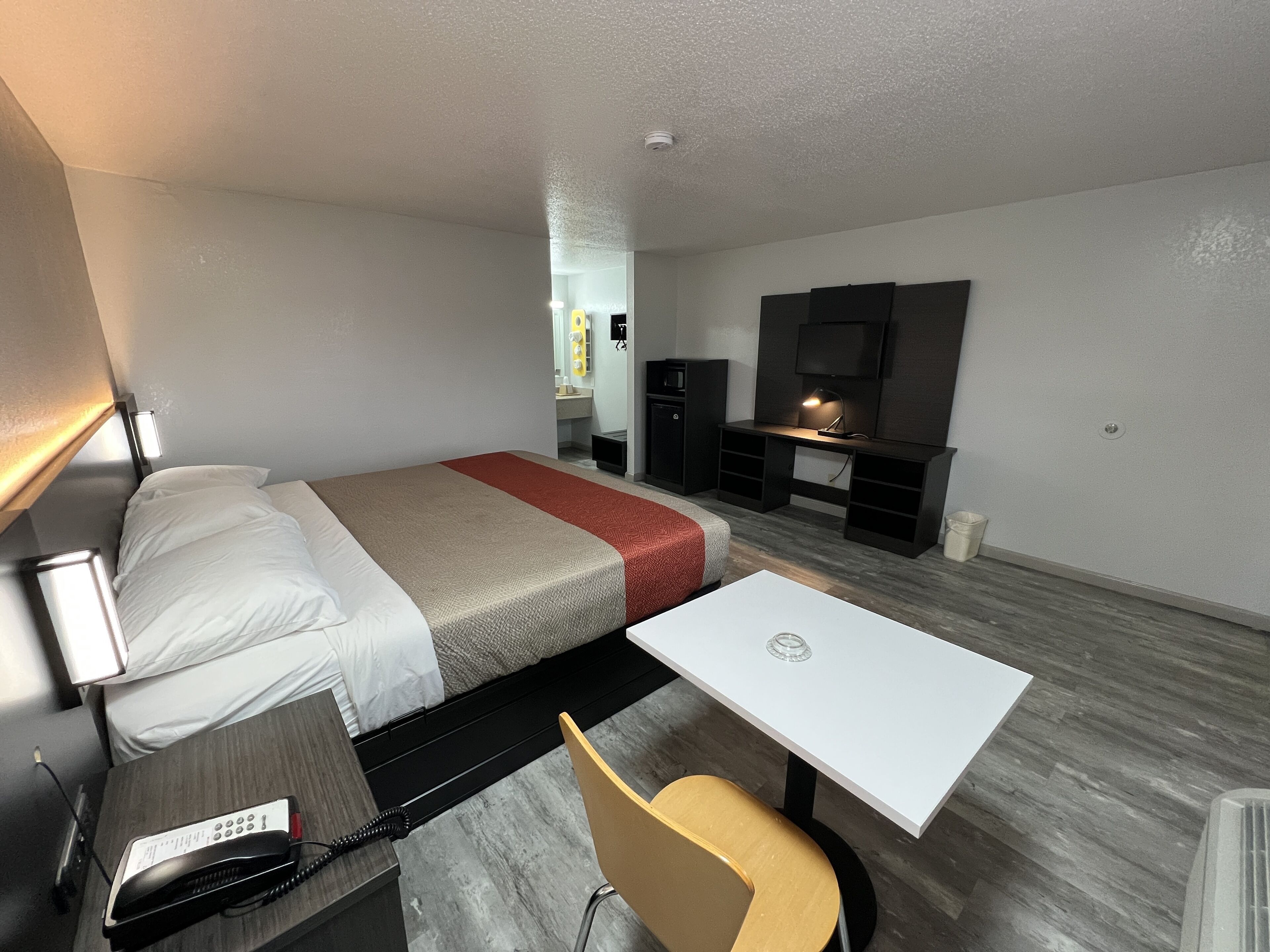 Quarto de hotel com cama, mesa de cabeceira, escrivaninha, cadeira e televisão.