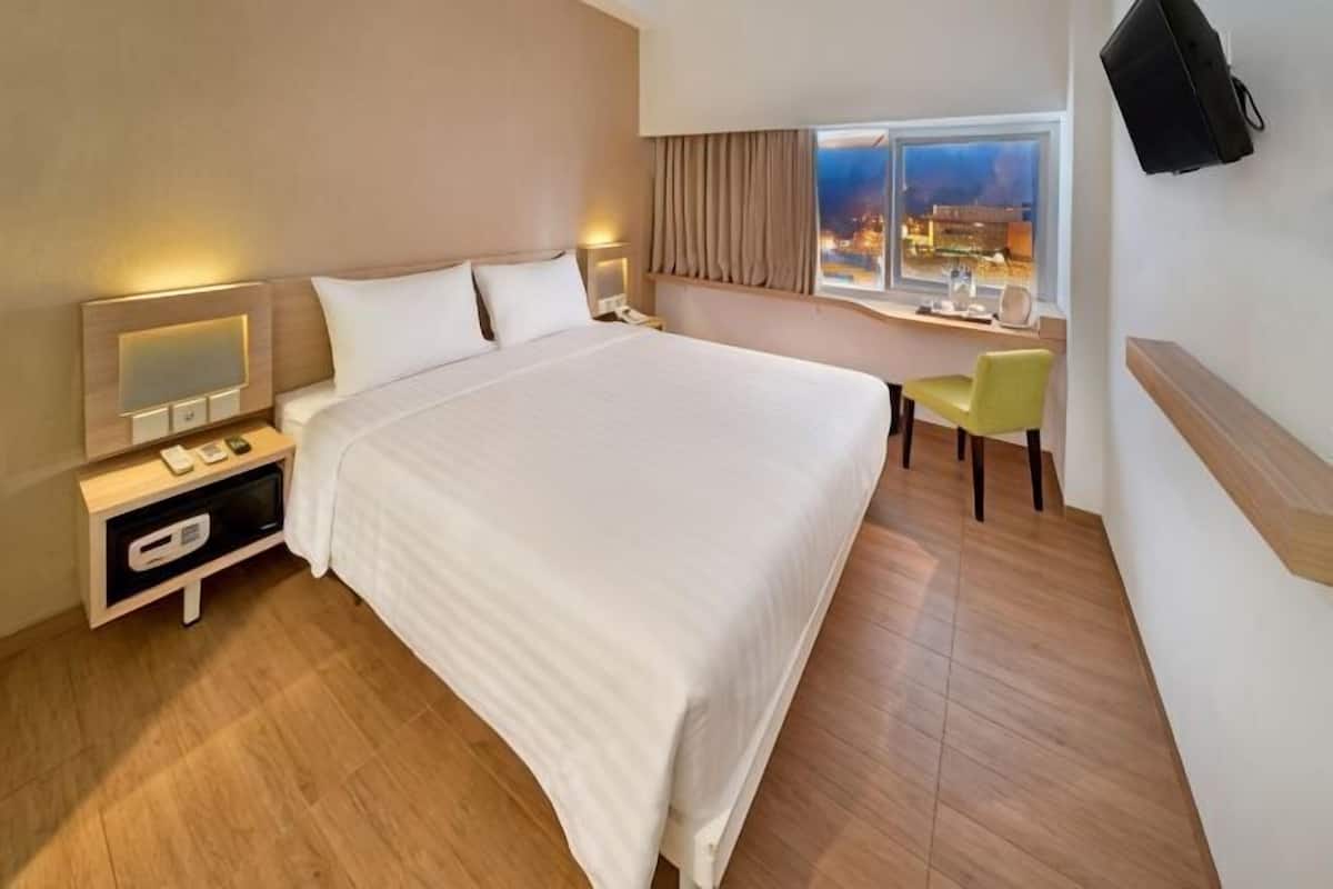 Quarto de hotel com uma cama grande, uma escrivaninha com cadeira, uma TV na parede e uma janela com vista para a cidade.