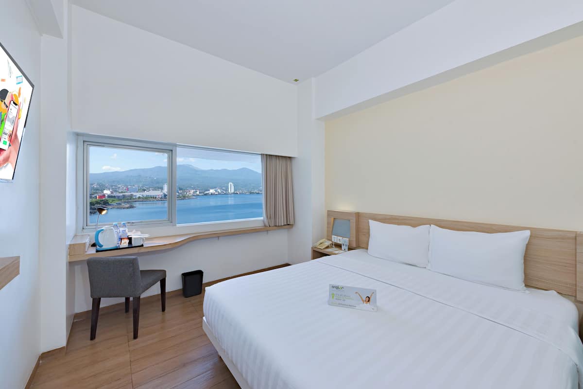 Quarto de hotel com uma cama grande, uma escrivaninha com cadeira, uma janela com vista para um corpo de água e montanhas, e uma mesa de cabeceira com abajur.