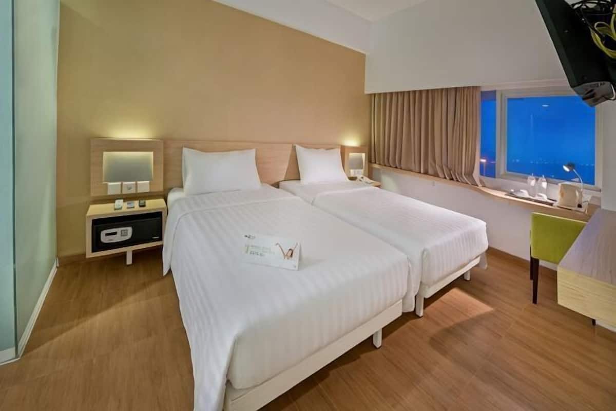Quarto de hotel com duas camas, uma escrivaninha, uma cadeira, uma mesinha de cabeceira e uma janela com vista.