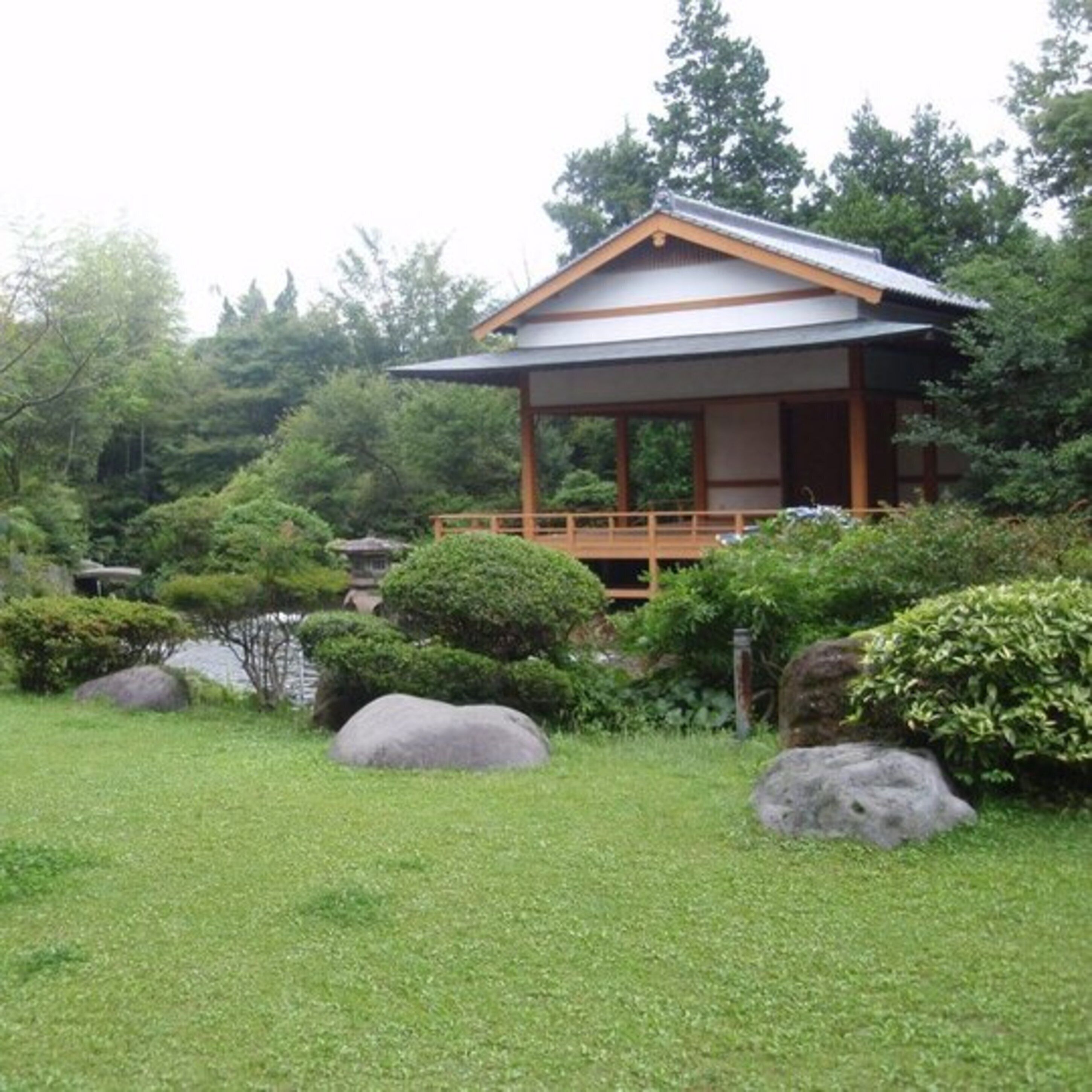 Um edifício tradicional em estilo japonês, com estrutura de madeira e telhado de telhas, cercado por vegetação exuberante e pedras.