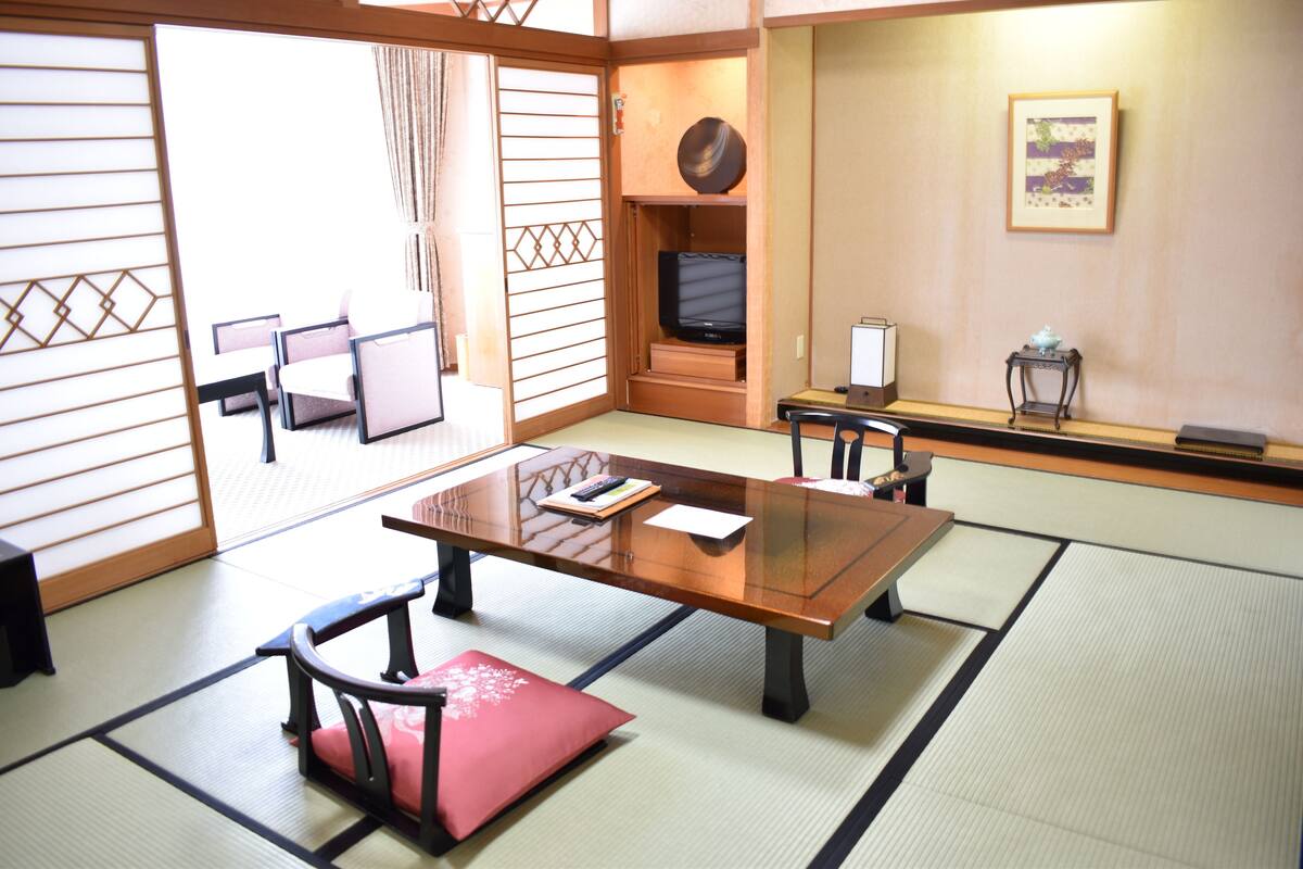 Um quarto japonês tradicional com tatames, uma mesa baixa com almofadas, uma televisão e um pequeno armário.