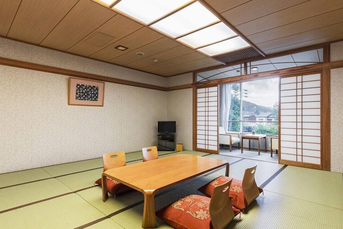 Um quarto japonês tradicional com tatames, uma mesa de madeira, cadeiras, uma televisão e portas deslizantes que dão para uma vista de montanha.
