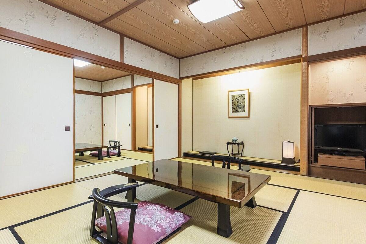 Um quarto japonês tradicional com tatames, uma mesa baixa, uma cadeira, uma televisão e um quadro na parede.