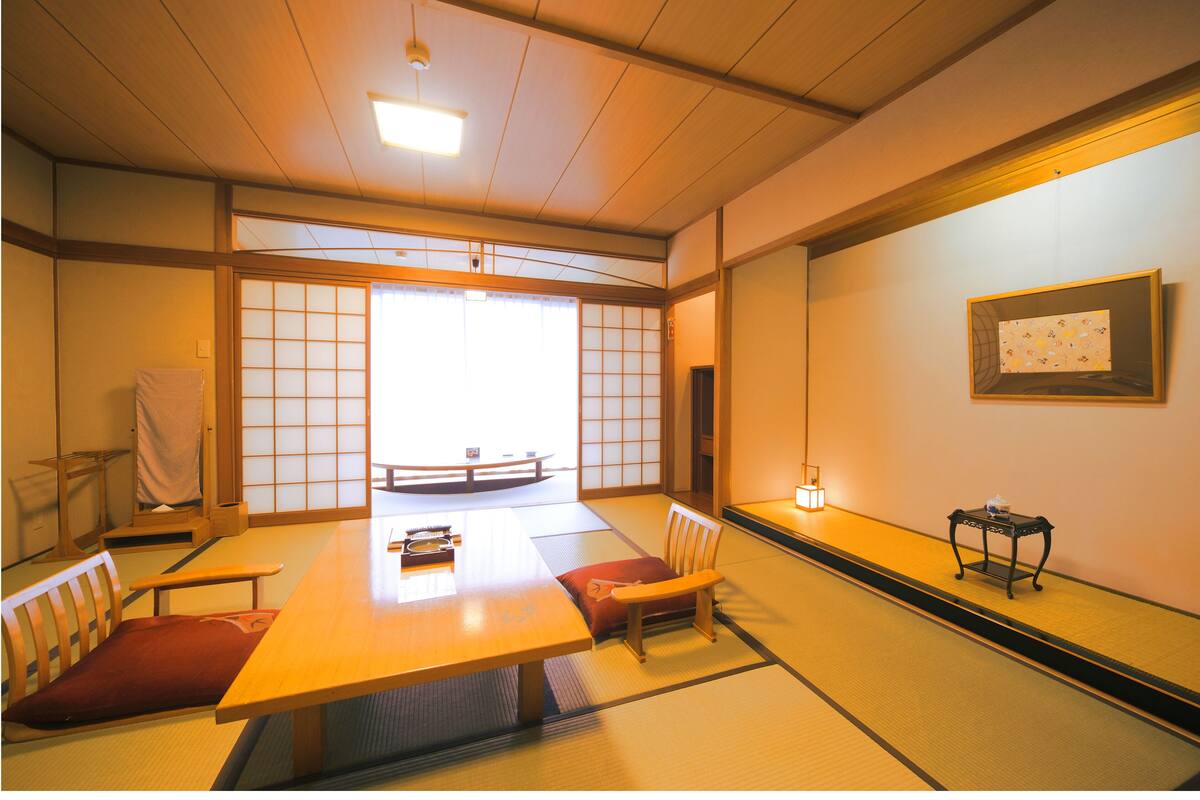 Um quarto japonês tradicional com tatames, uma mesa baixa, um banco acolchoado e uma porta deslizante para uma varanda.