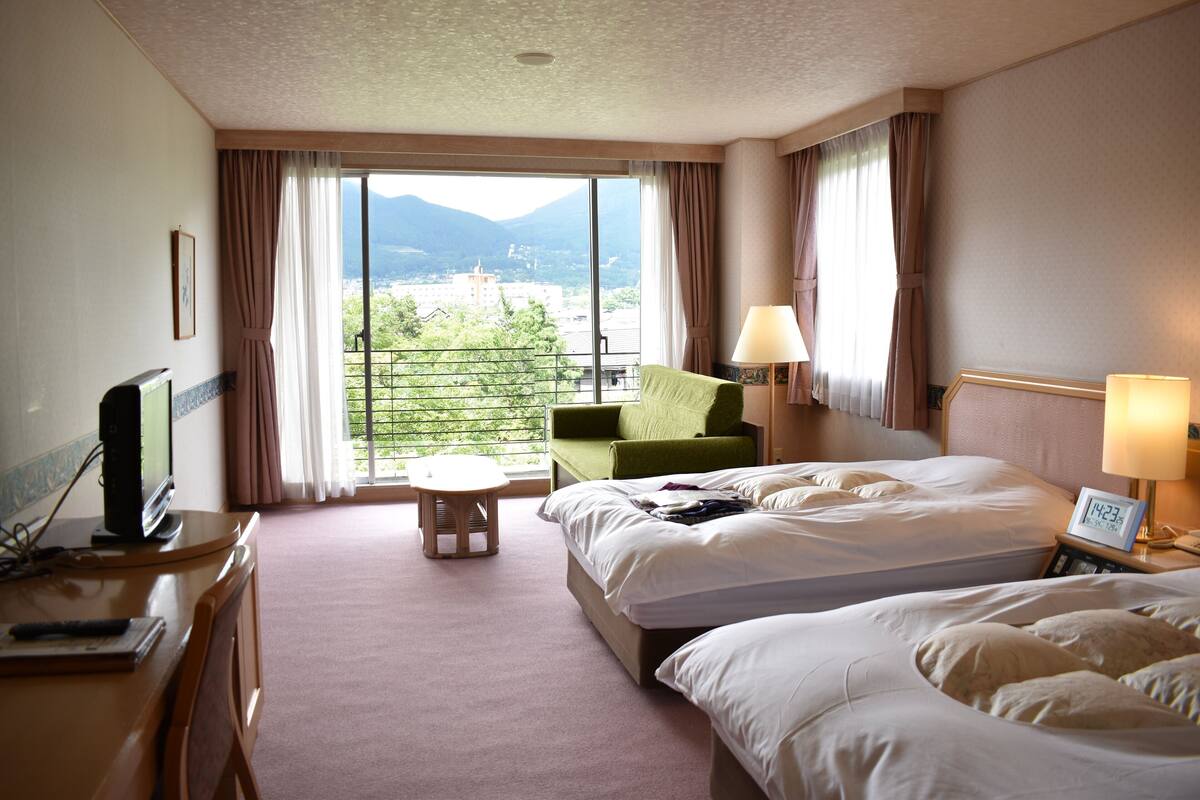 Quarto de hotel com duas camas, uma escrivaninha com computador, uma cadeira, uma mesa pequena e vista para montanhas pela janela.