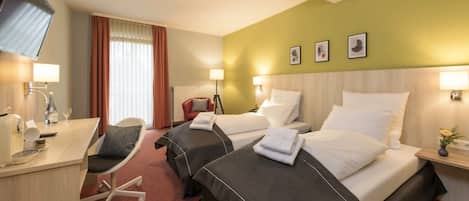 Quarto de hotel com duas camas, uma escrivaninha com cadeira, uma televisão e uma janela com cortinas.