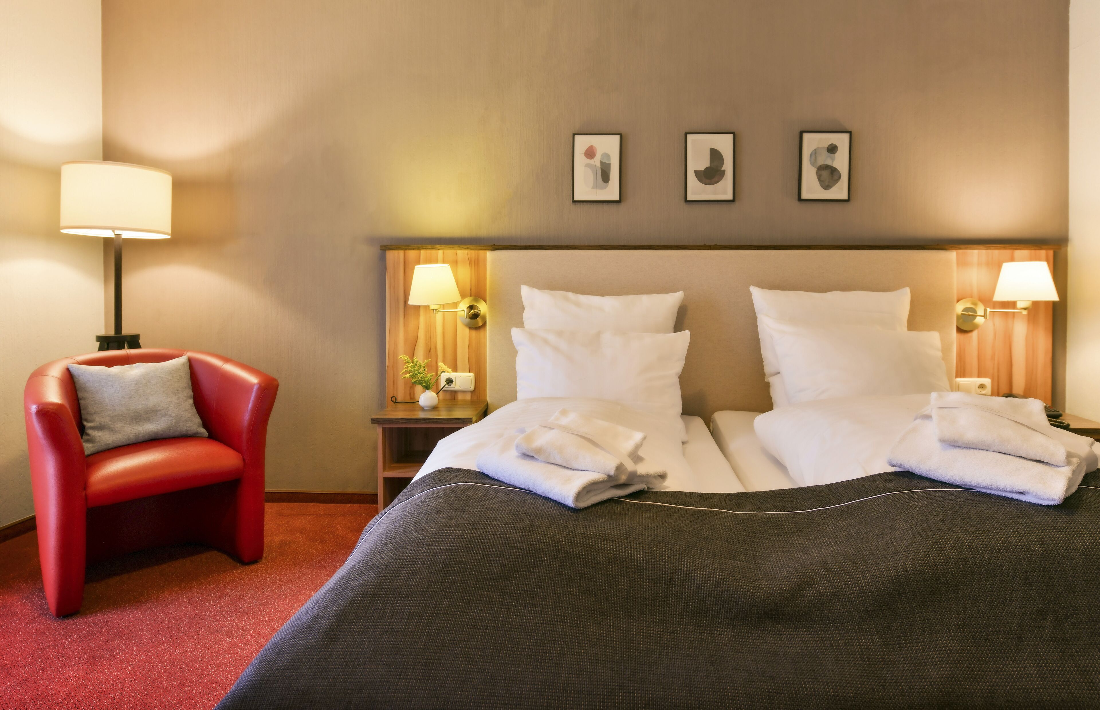 Quarto de hotel com cama, duas mesas de cabeceira com abajures, uma cadeira vermelha e obras de arte nas paredes.