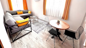 Uma sala de estar moderna com um sofá cinzento, almofadas amarelas, uma pequena mesa e um espaço de refeições com uma mesa redonda e cadeiras.