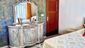 Quarto com cômoda de estilo vintage, um espelho, porta vermelha e cama com colcha florida.