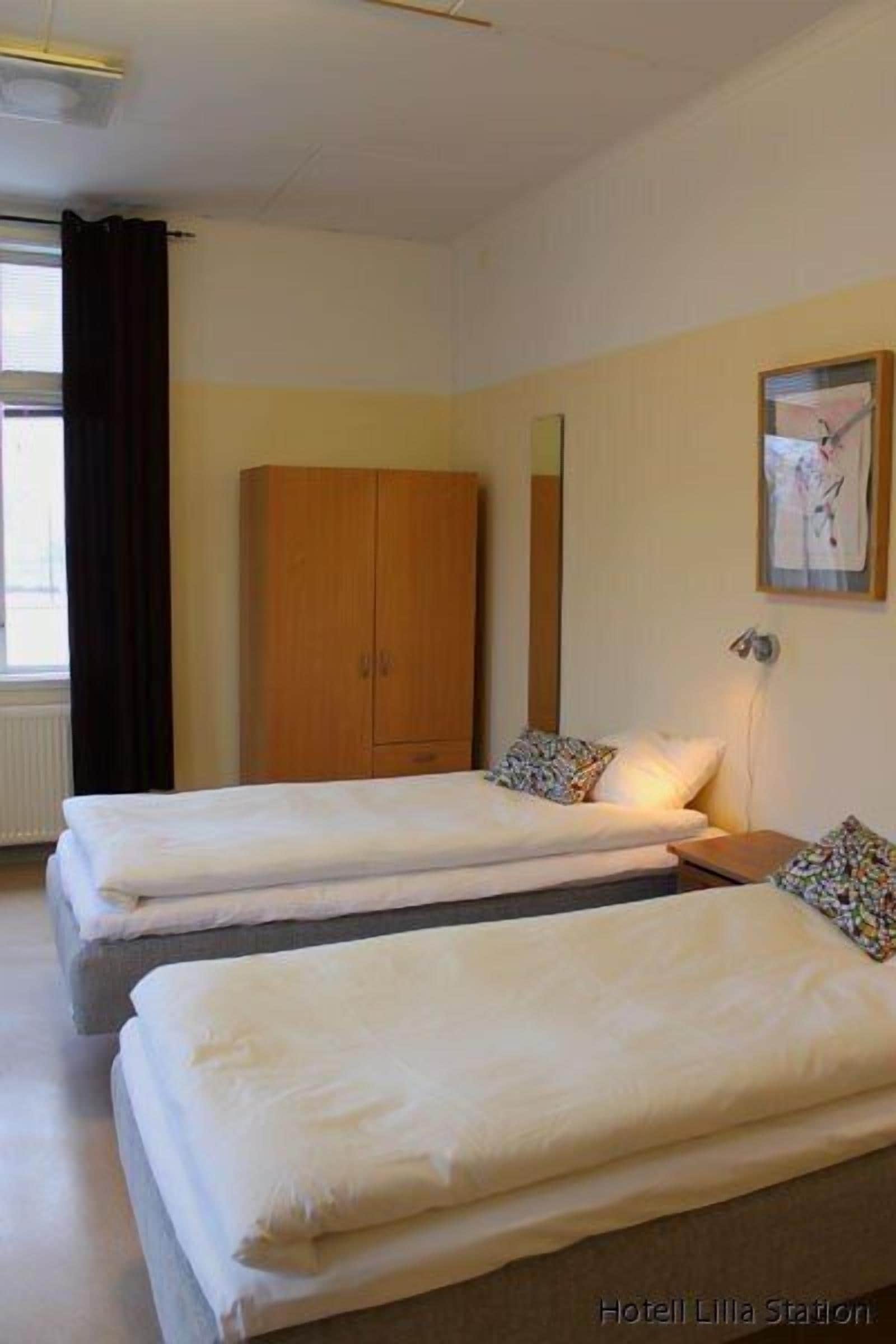 Quarto de hotel com duas camas, um guarda-roupa, uma janela com cortinas e uma luminária fixada na parede.