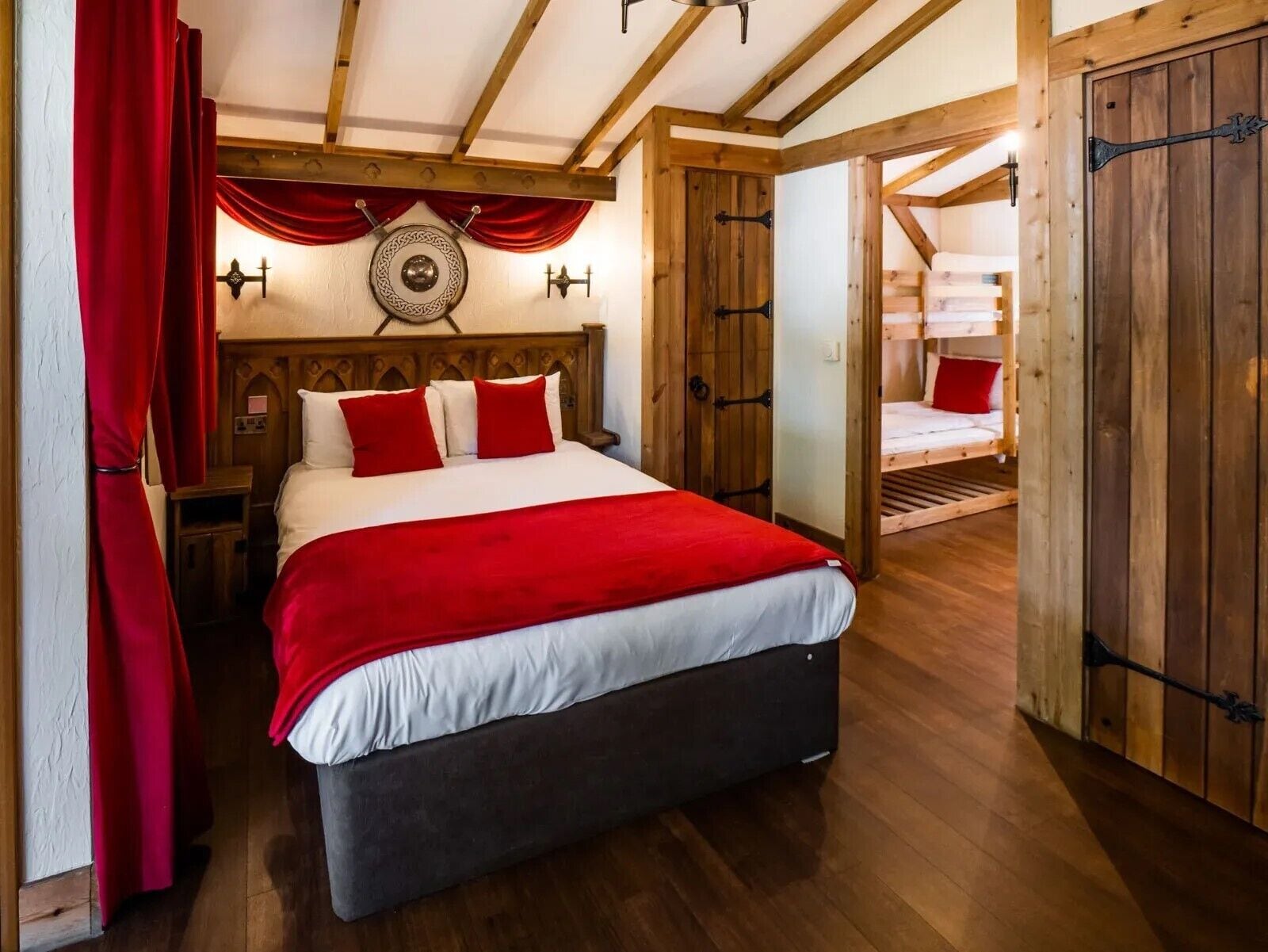 Quarto com cama grande, vigas de madeira, cortina vermelha e porta com puxador de metal.