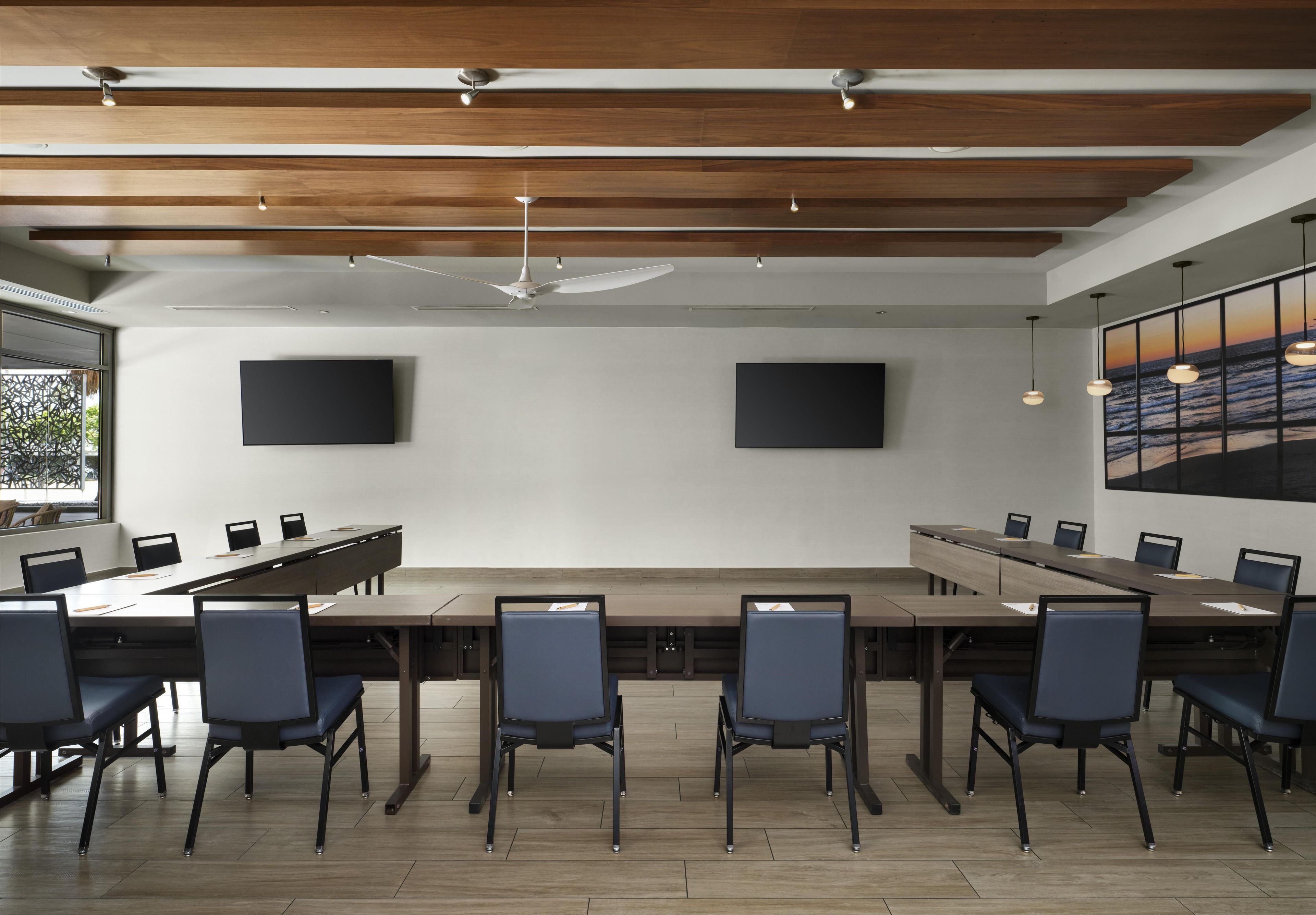 Uma sala de conferência com uma mesa comprida, cadeiras, ventiladores de teto e duas televisões de tela plana fixadas nas paredes.