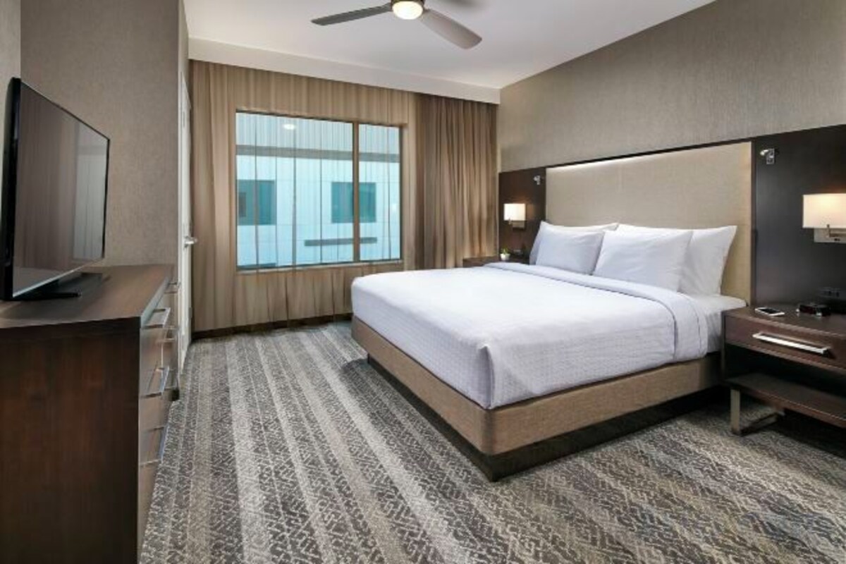 Quarto de hotel com cama, televisão, secretária e janela com cortinas.