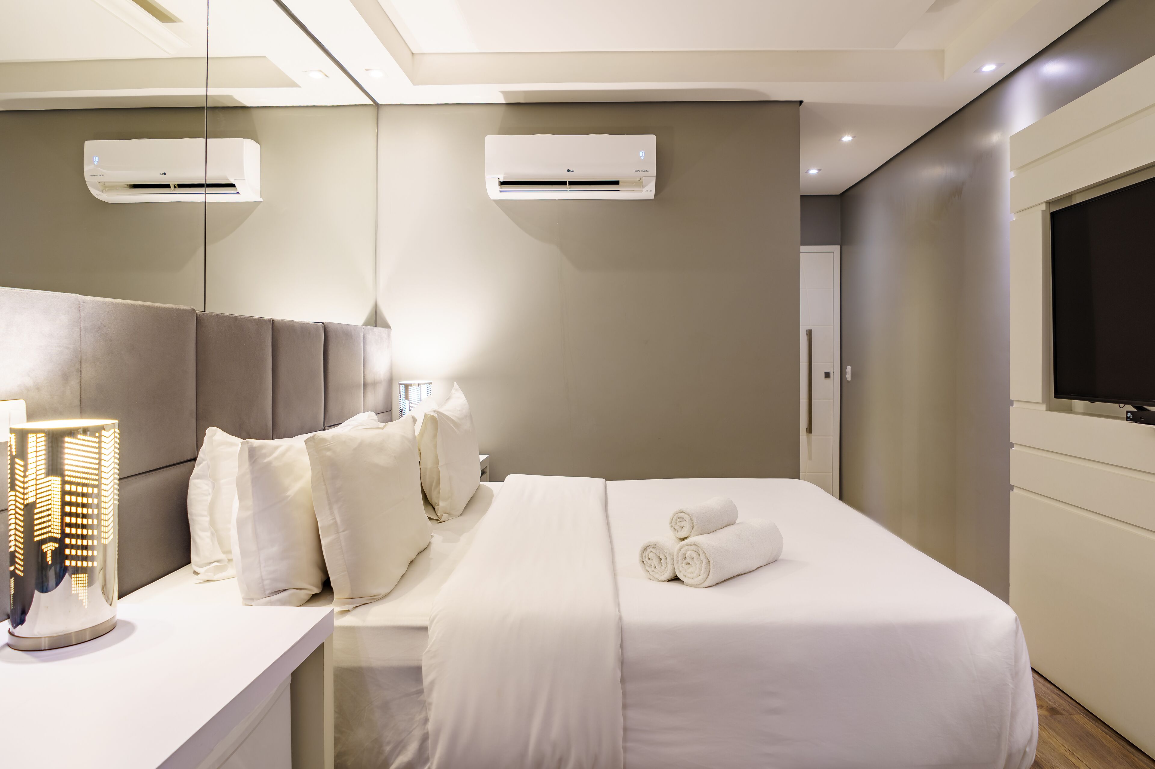 Quarto de hotel moderno com uma cama grande, uma mesa de cabeceira com abajur e duas unidades de ar condicionado na parede.