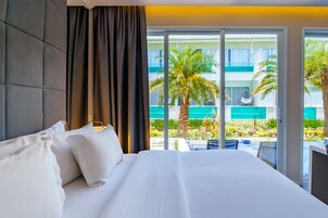 Quarto de hotel com uma cama grande, vista para uma piscina e palmeiras, e varanda com mesa e cadeiras.