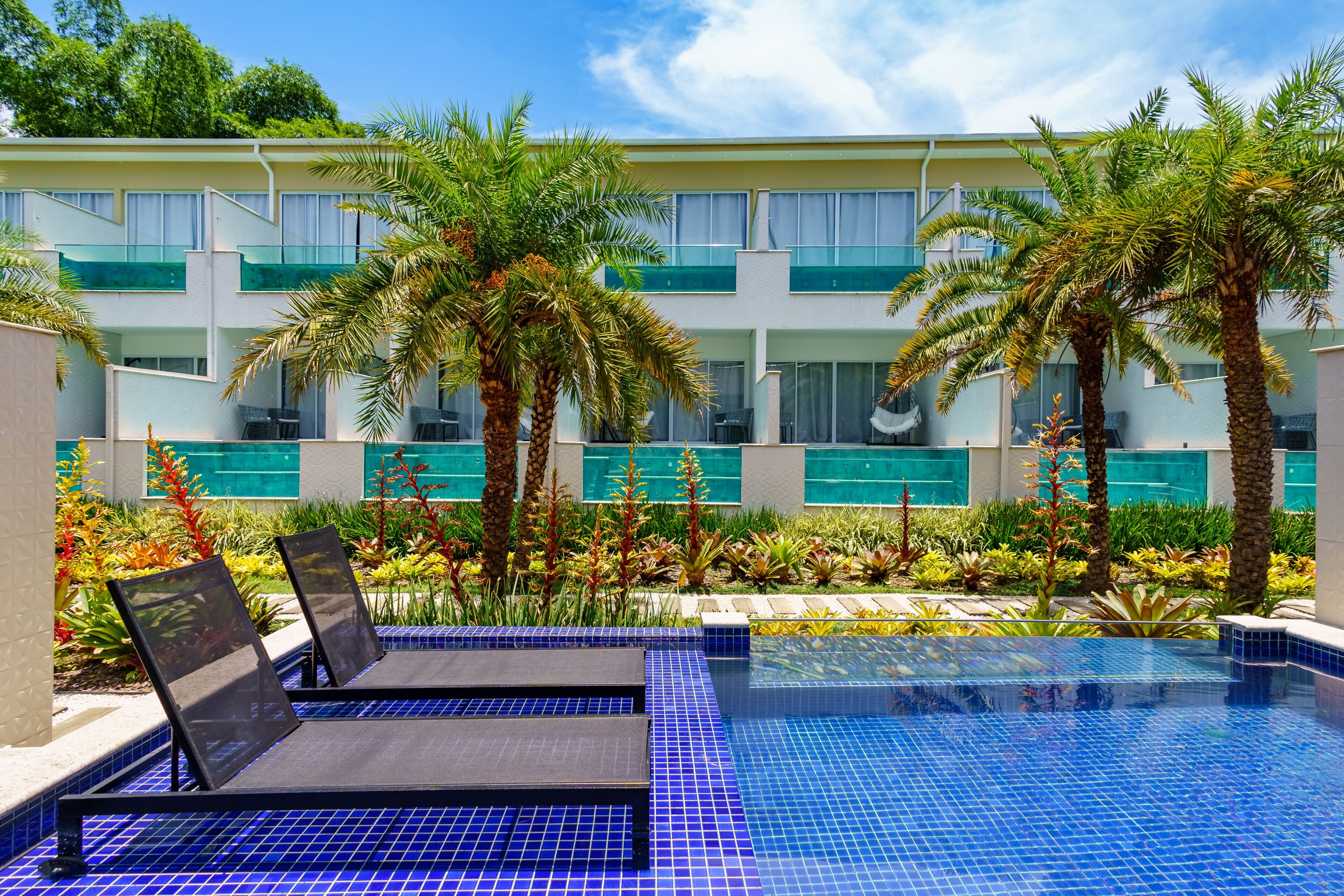 Uma área de piscina com espreguiçadeiras, palmeiras e um edifício com varandas.