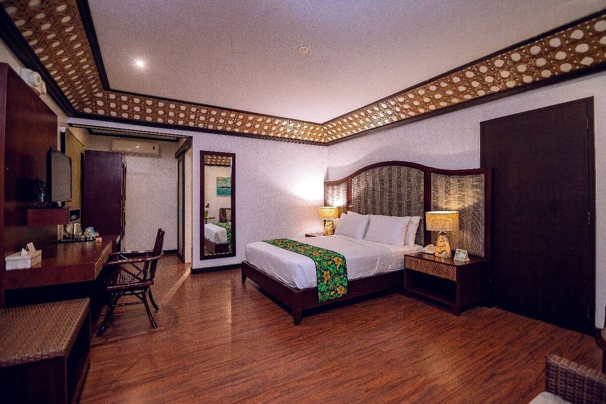 Quarto de hotel com soalho de madeira, cama com colcha floral verde, mesa de cabeceira com abajur, guarda-roupa de madeira e espelho na parede.