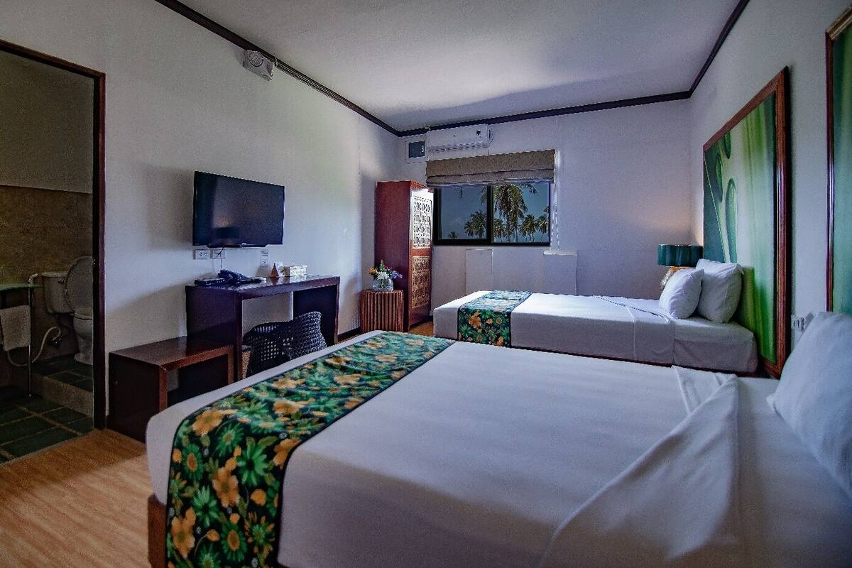 Quarto de hotel com duas camas, uma mesa de trabalho com televisão, janela com vista para palmeiras e uma mesinha com um vaso de flores.