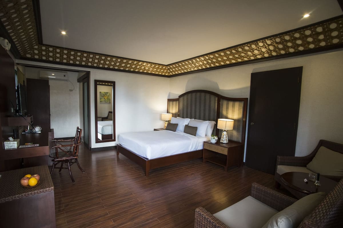 Quarto de hotel com uma cama grande, duas mesas de cabeceira com abajures, uma cadeira, uma mesinha com um copo e uma porta.