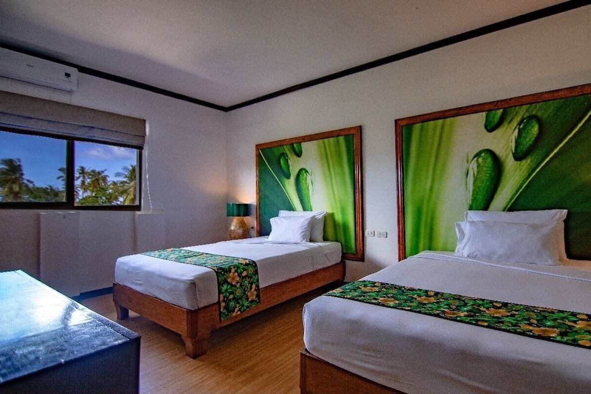 Quarto de hotel com duas camas de solteiro, uma mesinha e janela com vista para palmeiras.