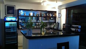 Uma área de bar com geladeira repleta de garrafas, prateleiras com copos e um vaso de planta no balcão.