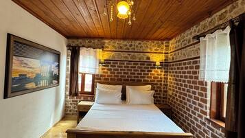 Teto de madeira com luminária pendente, cama com roupa de cama branca, parede de pedra e um quadro na parede.