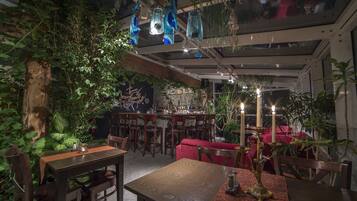 Espaço de refeições interno com mesas e cadeiras de madeira, um balcão com banquetas e garrafas de vidro penduradas.
