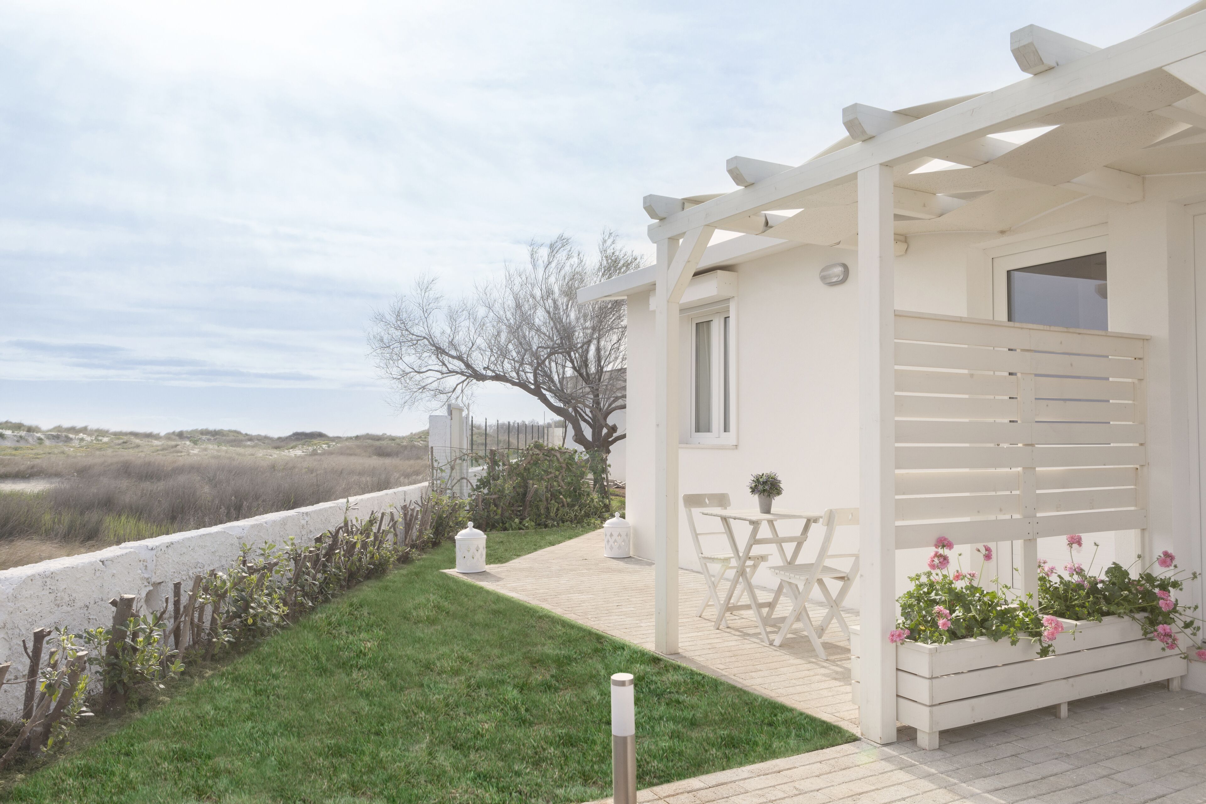 Um pátio externo com piso branco, uma pérgola, uma mesa e cadeiras, e um vaso com flores.