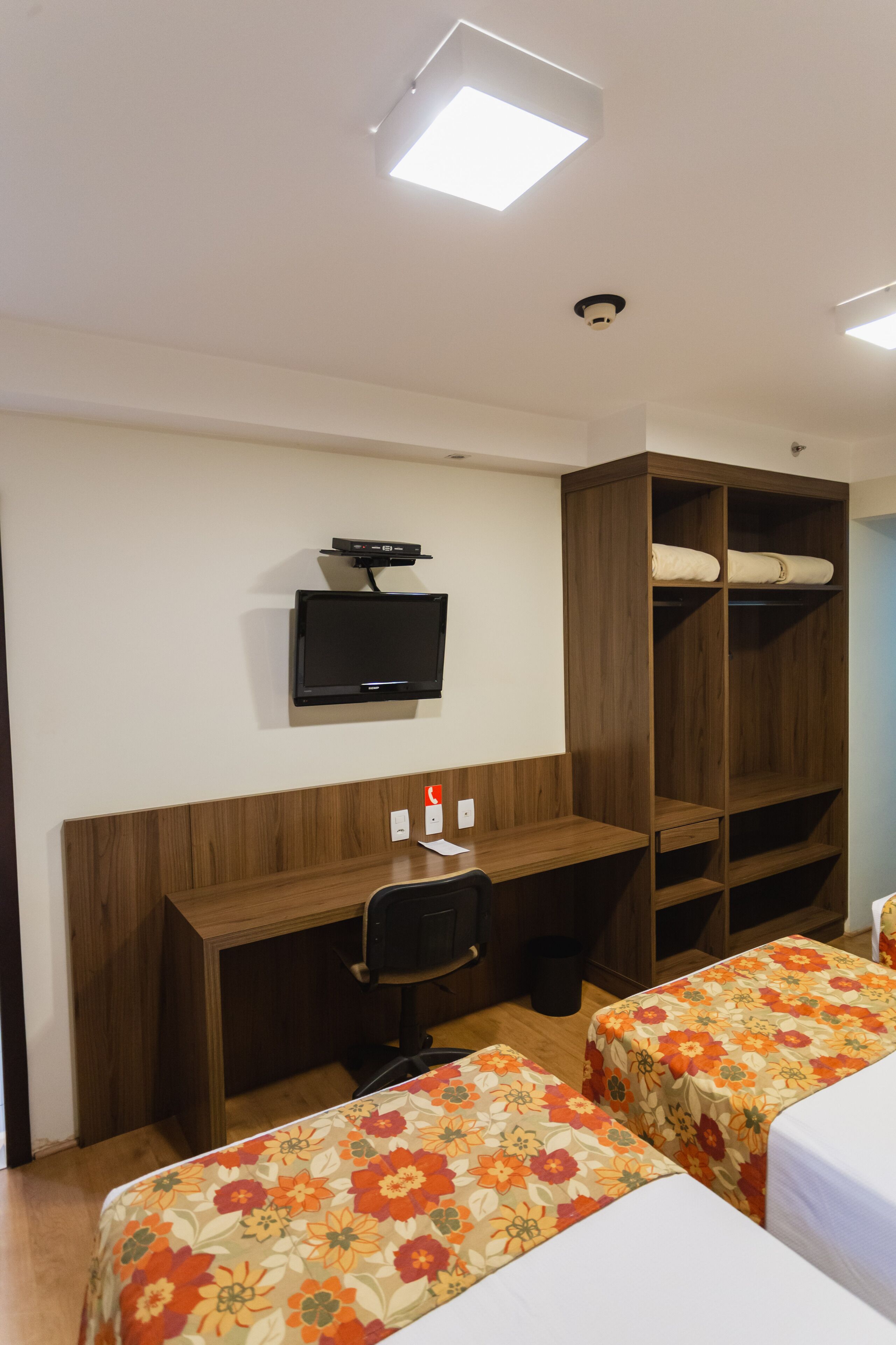Quarto de hotel com escrivaninha de madeira, televisão fixada na parede, beliche com cabeceira de madeira e duas camas com colchas floridas.