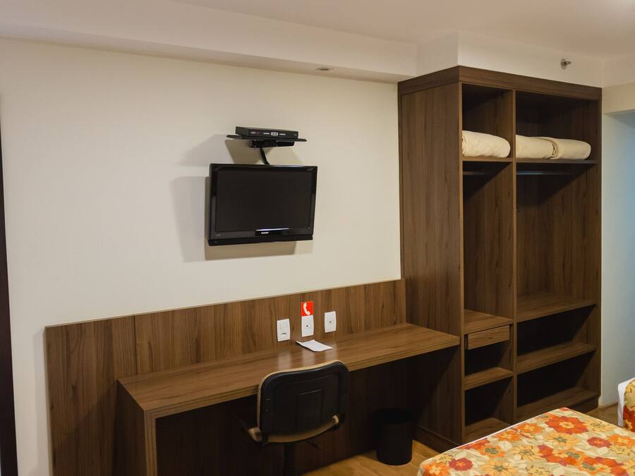 Quarto de hotel com escrivaninha de madeira, televisão fixada na parede, beliche com cabeceira de madeira e duas camas com colchas floridas.