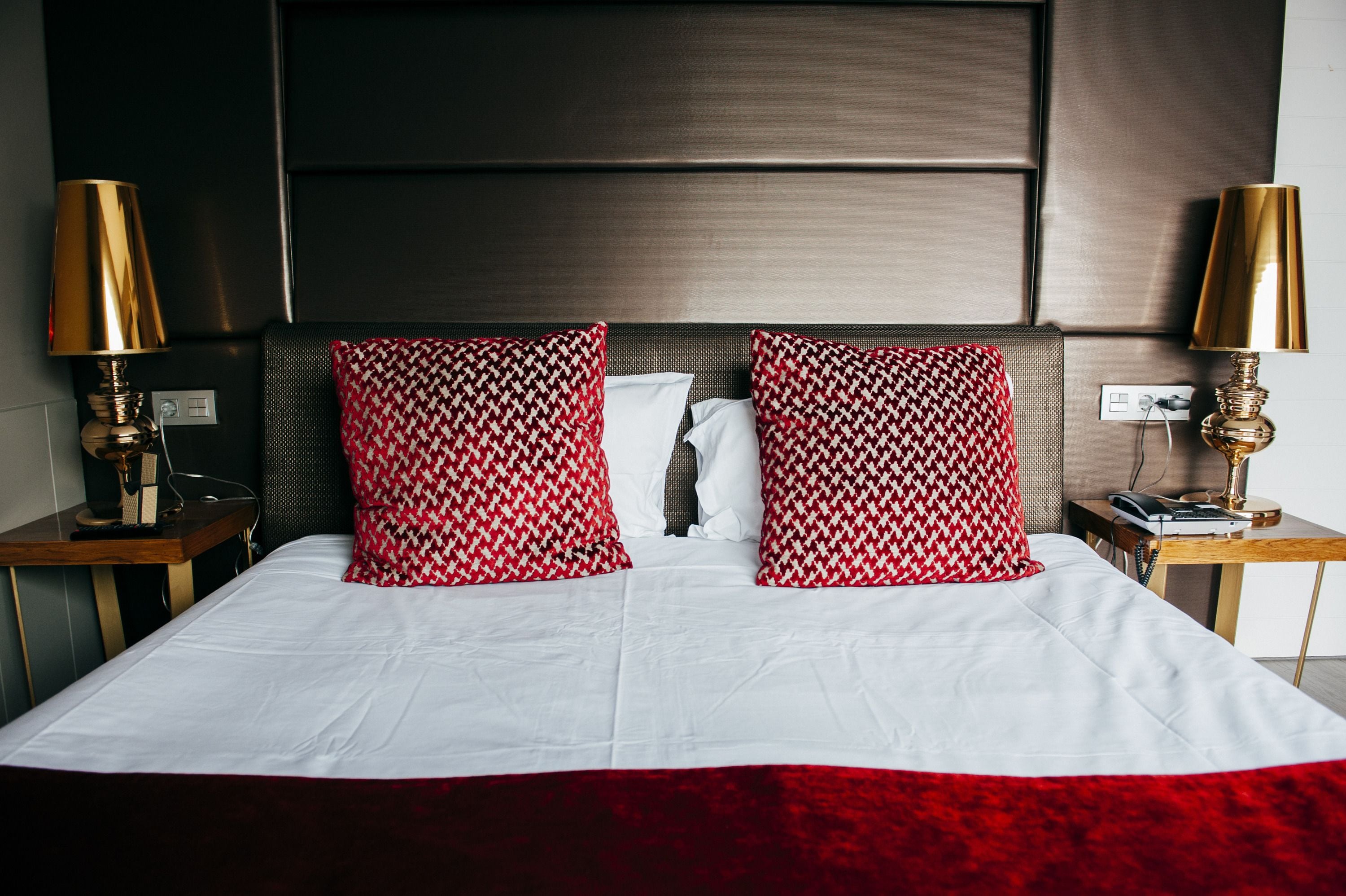 Uma cama bem arrumada com lençóis brancos e almofadas vermelhas estampadas, duas luminárias de cabeceira e um cobertor vermelho.
