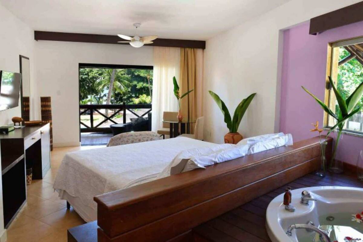 Quarto de hotel com cama, banheiro com banheira, escrivaninha com cadeira e vista para o verde lá fora.
