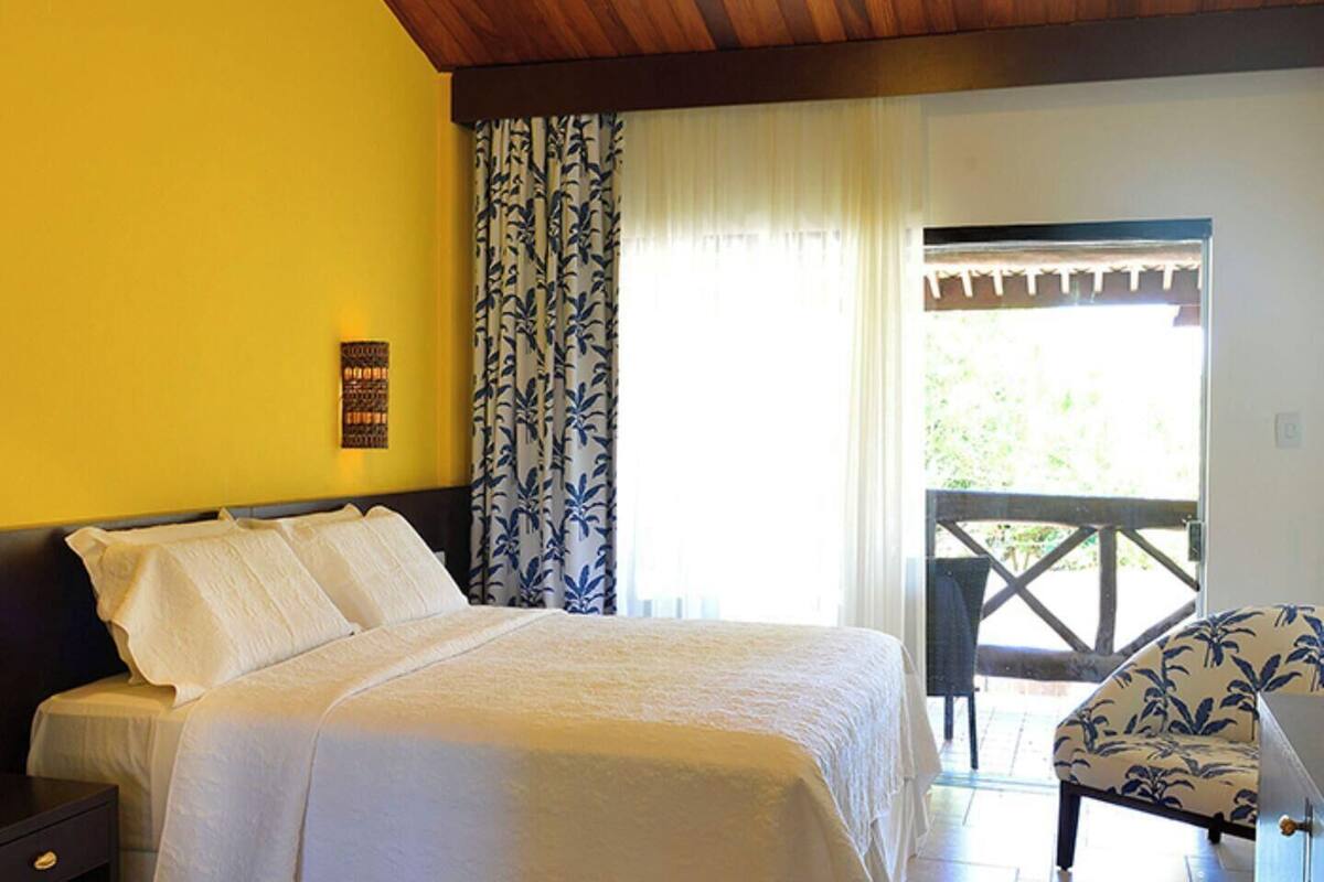 Quarto de hotel com teto de madeira, cama com roupa de cama branca, cortina estampada e vista para o exterior.