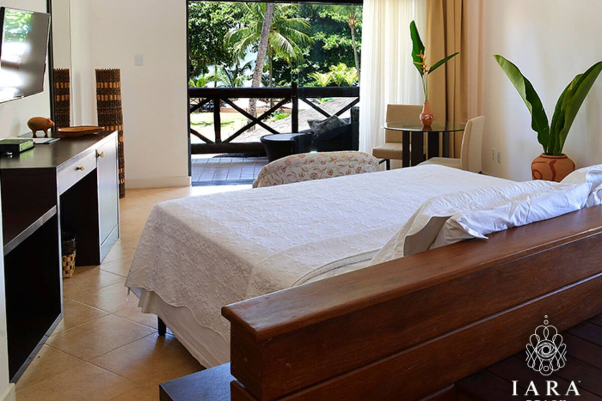Quarto de hotel com cama, escrivaninha, televisão, ventoinha no teto e vista para o exterior.