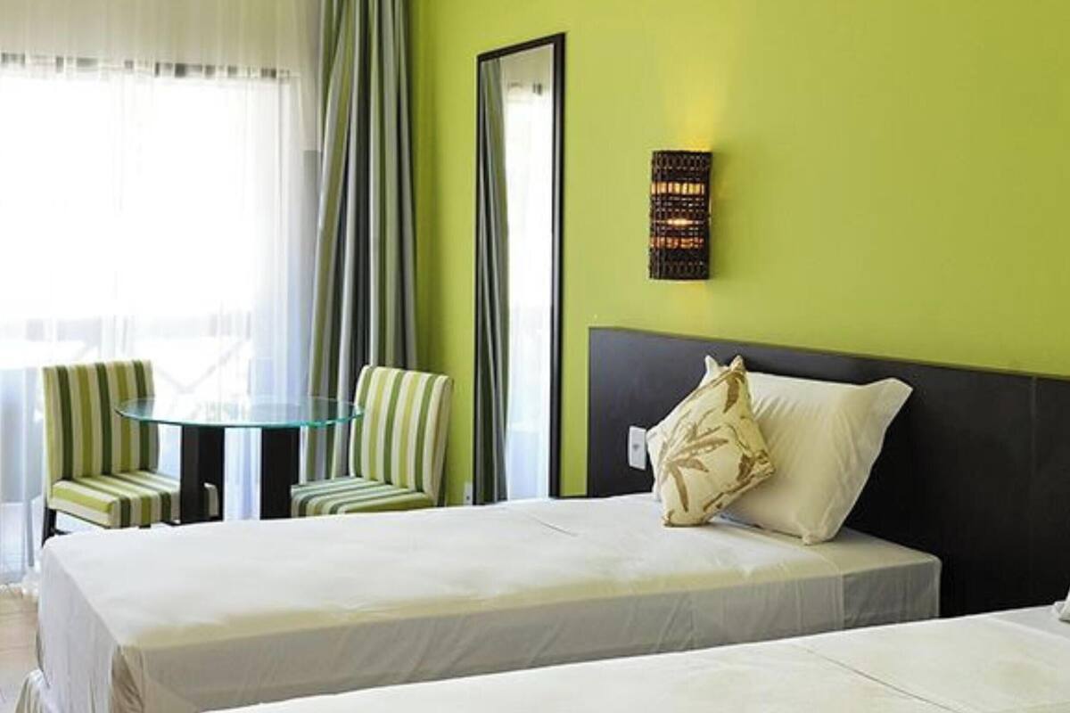 Quarto de hotel com duas camas, uma mesa de jantar, cadeiras, um ventilador de teto, um espelho e um luminário fixado na parede.