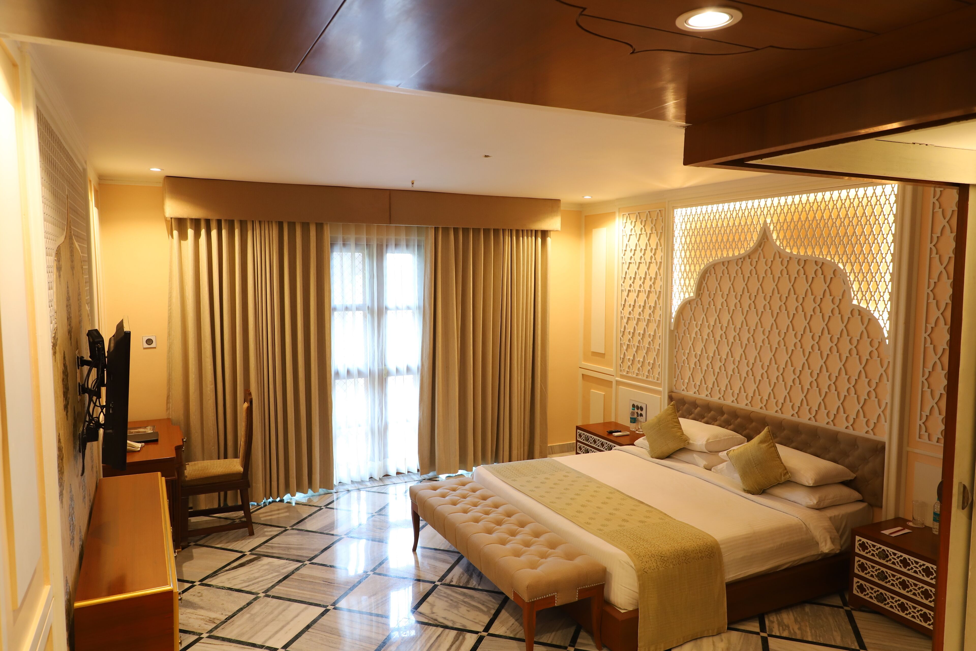Quarto de hotel bem iluminado com uma cama grande, um banco, uma escrivaninha e uma cadeira.