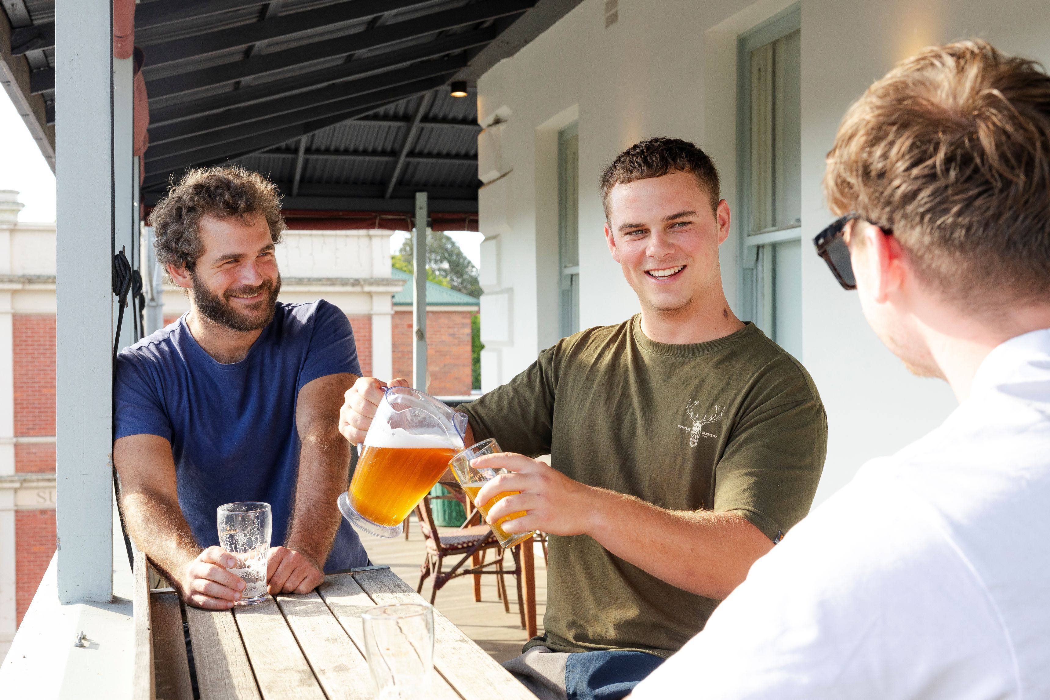 Três pessoas brindando com copos de cerveja em uma mesa de madeira sob um toldo.