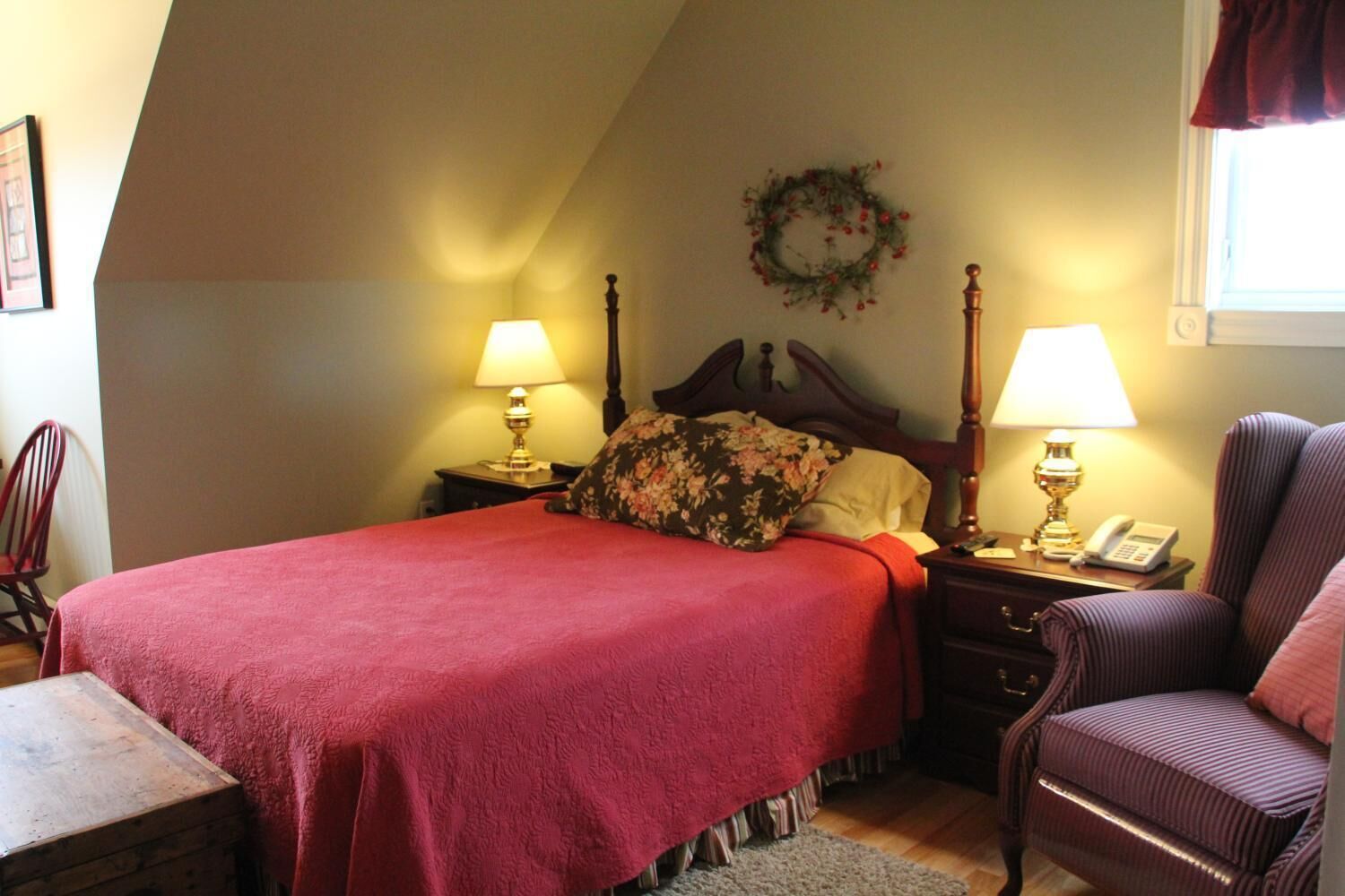 Quarto com cama grande, duas mesas de cabeceira com abajures, uma cadeira, um baú de madeira e uma janela com cortinas.