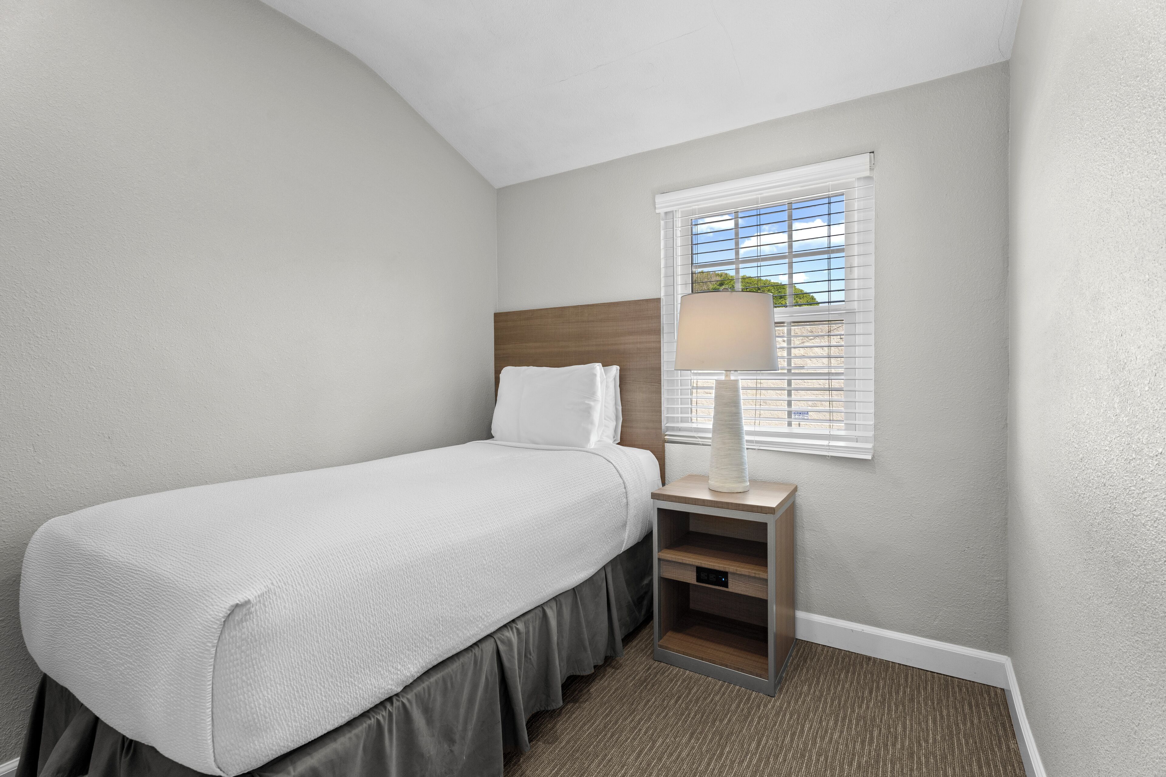 Quarto de hotel bem arrumado, com uma cama de solteiro, mesa de cabeceira com abajur e janela com persianas.