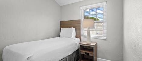 Quarto de hotel bem arrumado, com uma cama de solteiro, mesa de cabeceira com abajur e janela com persianas.