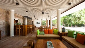 Um espaço interno amplo com piso de madeira, um longo banco de madeira e um sofá com almofadas coloridas. Há várias mesas e cadeiras, e uma grande janela que oferece vista para o exterior.