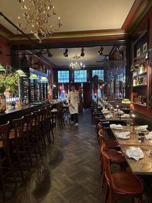 Interior de restaurante com bar, mesas de jantar, cadeiras e um candelabro.