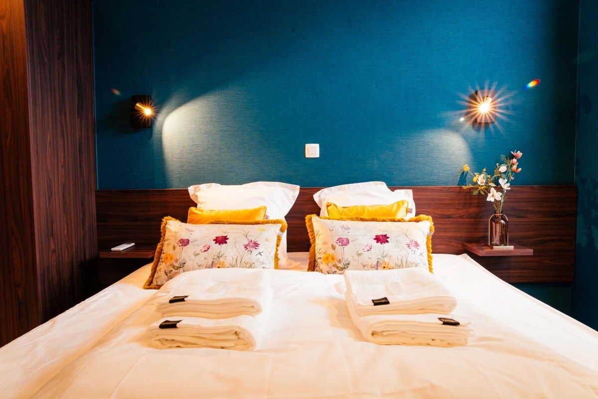 Quarto de hotel com uma cama grande, duas almofadas, mesa de cabeceira, um vaso com flores e uma luminária na parede.