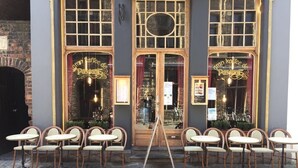 Um café com fachada de tijolos, grandes janelas e área externa com cadeiras de vime.