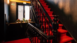 Uma escadaria com carpete vermelho e balaústres pretos, que leva a um quarto com parede escura e quadros emoldurados.