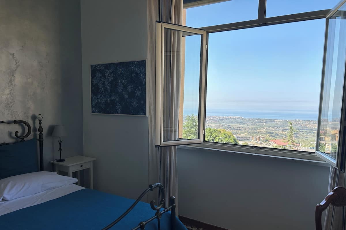 Quarto com cama, mesa de cabeceira, abajur e janela com vista para o mar e paisagem.