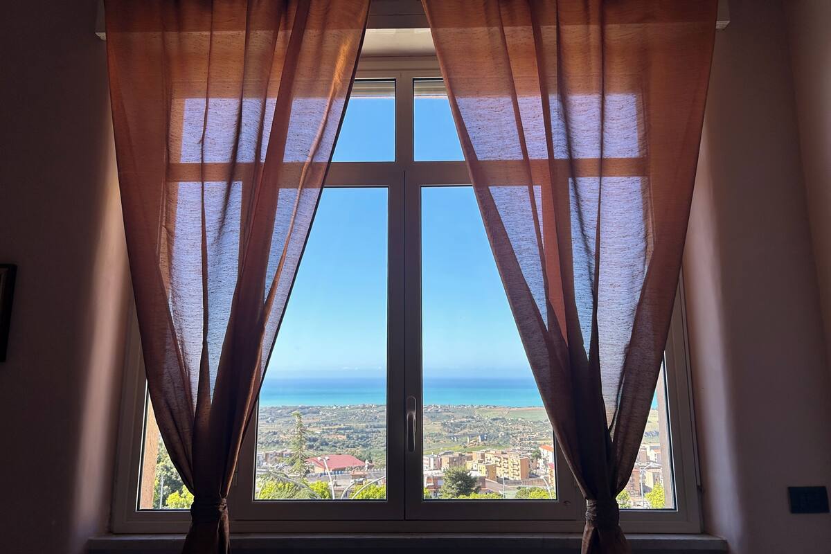 Janela com cortinas leves, exibindo vista de paisagem costeira com edifícios e vegetação.